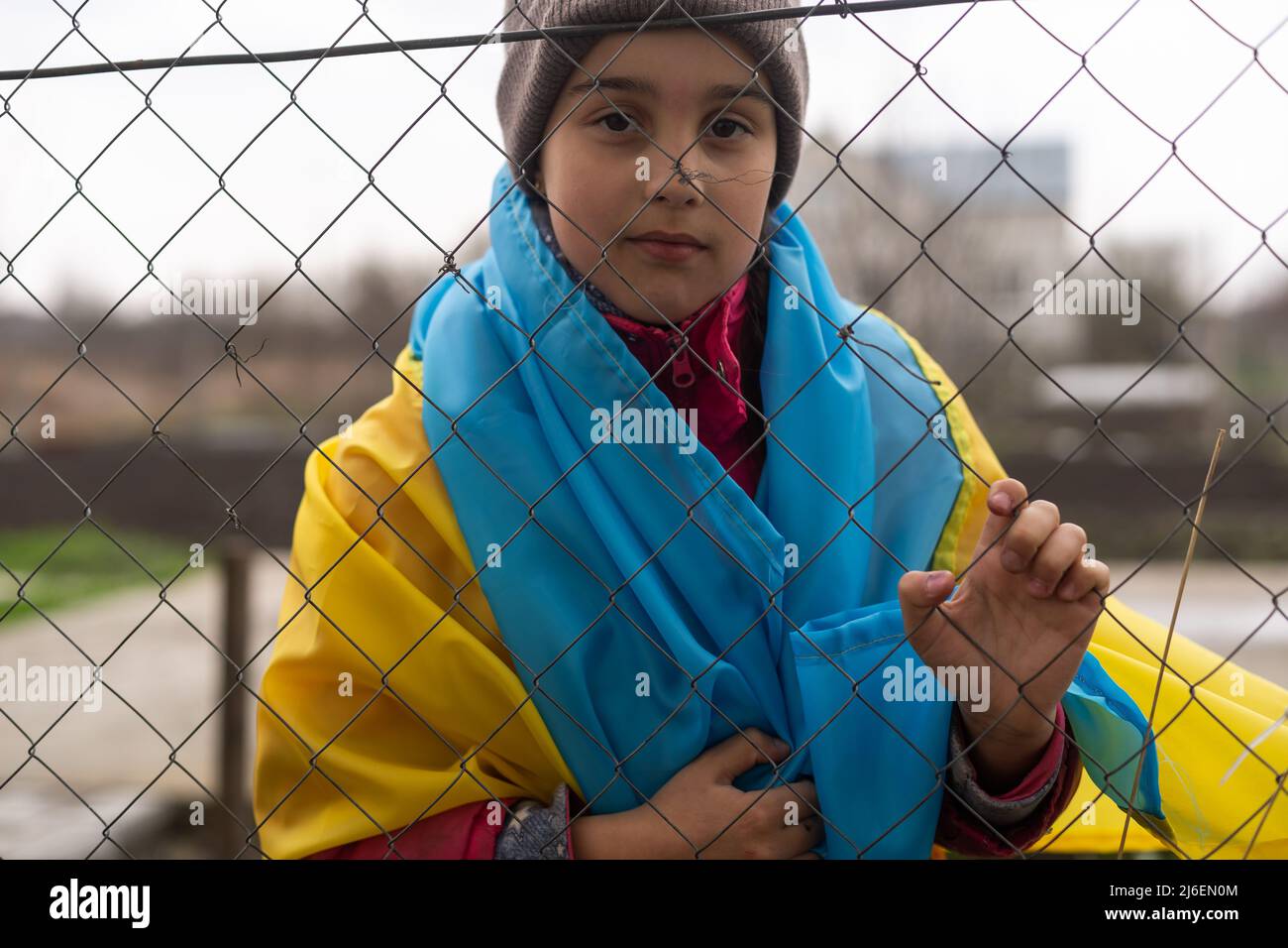 sad little girl with the flag of Ukraine behind a metal fence. Social ...