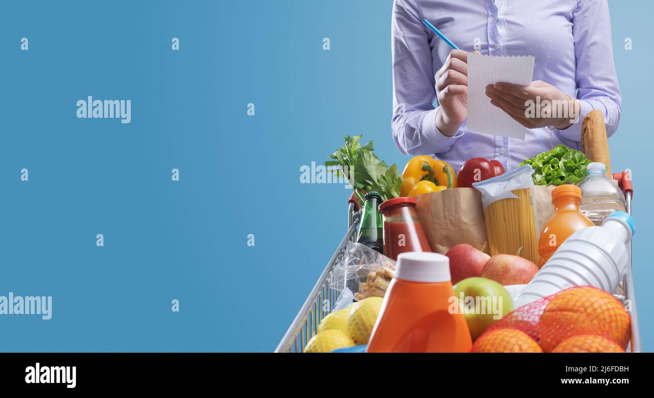 Woman writing a shopping list and trolley full of fresh goods, grocery ...