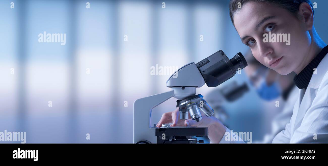 Young female researcher using a microscope and looking at camera ...