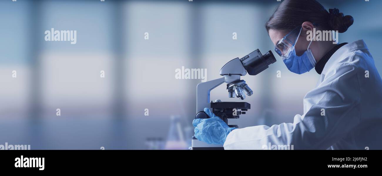 Researcher wearing a surgical mask and looking through a microscope ...