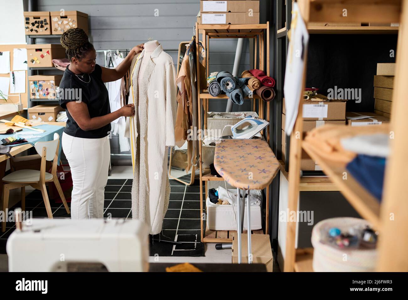 Contemporary seamstress taking measures of unfinished coat of pastel color hanging on mannequin ...