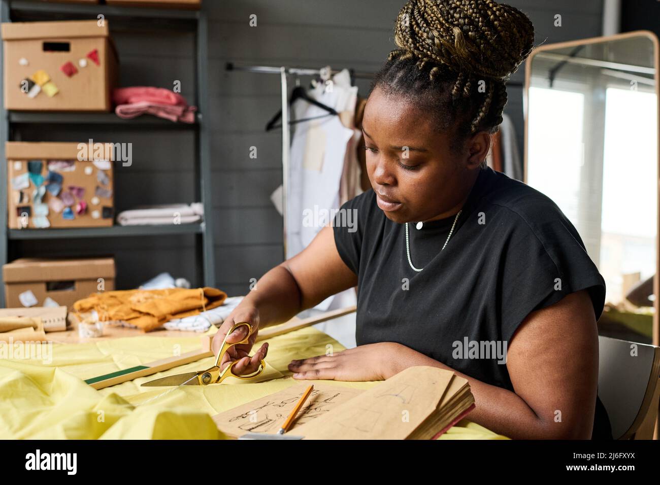 Young serious seamstress with scissors cutting textile of yellow color ...