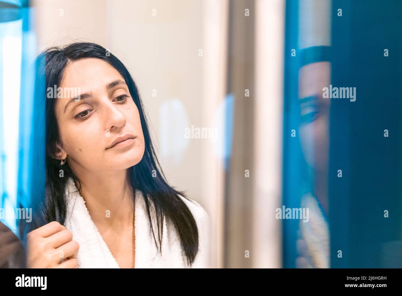 Pretty woman with fresh morning face in white robe looks to the mirror ...
