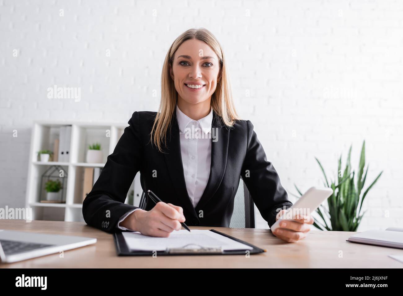 happy lawyer looking at camera while holding smartphone and pen near ...