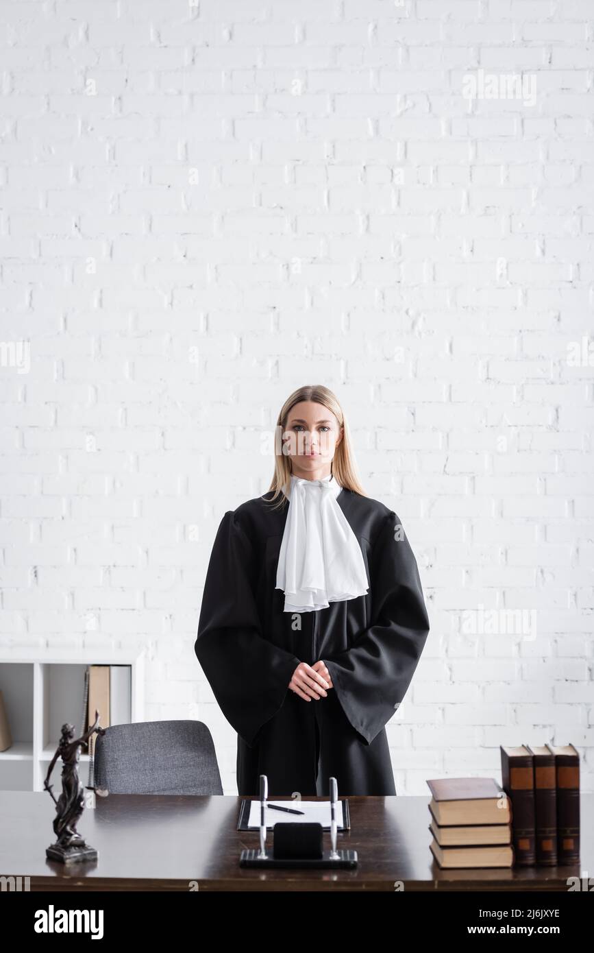 prosecutor in black mantle looking at camera near desk and white wall ...