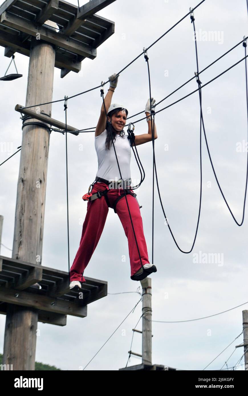 A young woman in a rope amusement park crosses a suspension bridge on a ...