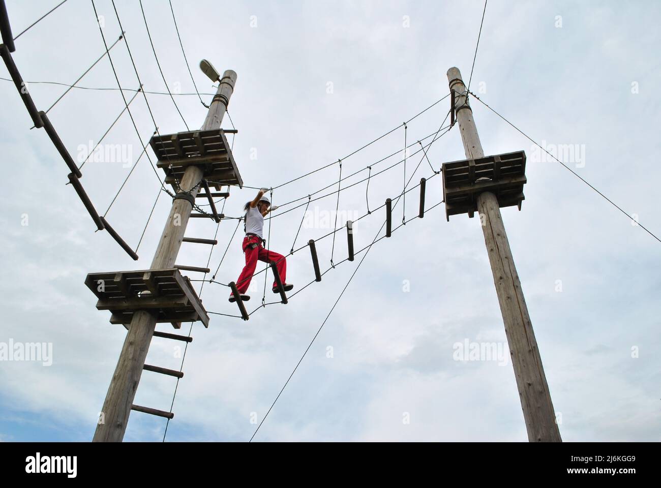 A young woman in a rope amusement park crosses a suspension wooden ...