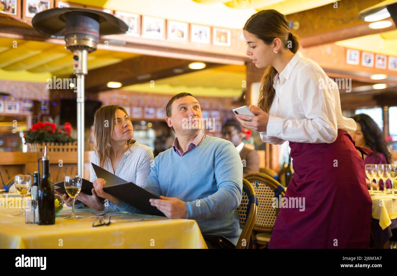 Girl waiter helps a couple choose dishes from the menu in restaurant ...