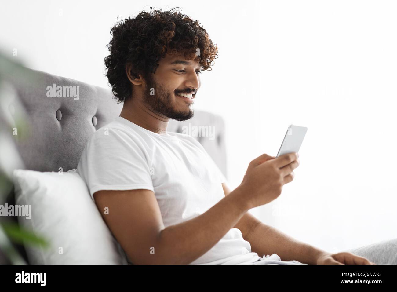 Positive indian guy scrolling in bed, using smartphone Stock Photo - Alamy