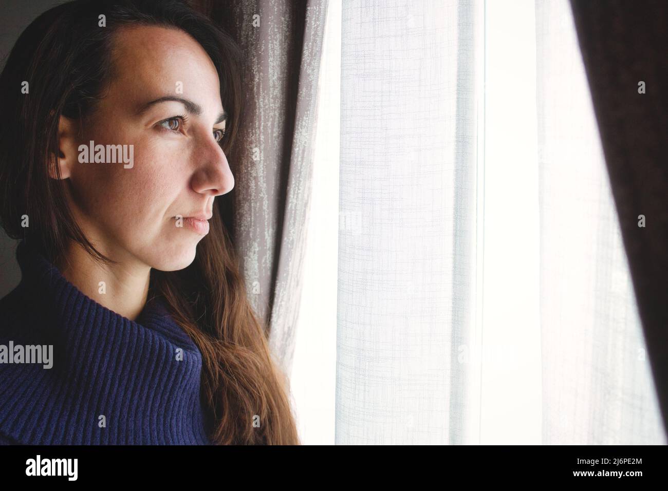 Young attractive woman looking pensively out of a window with curtain ...