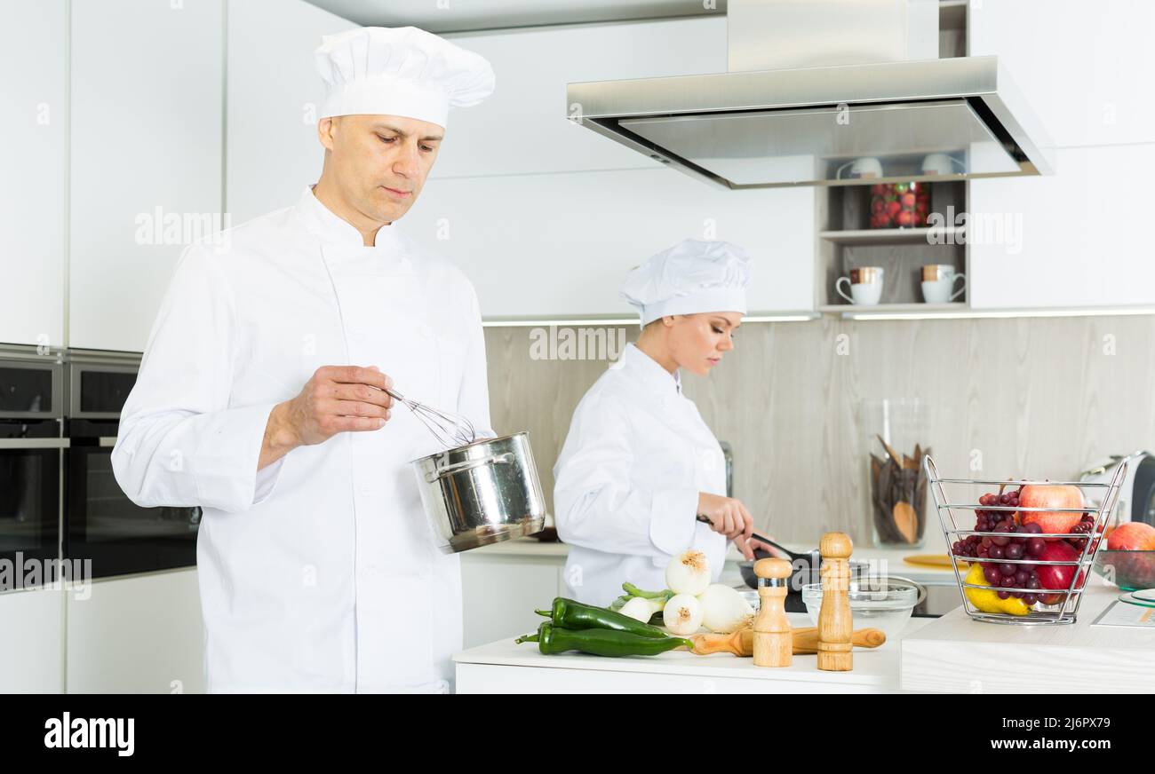Two positive female and male young cooks wearing uniform working Stock ...