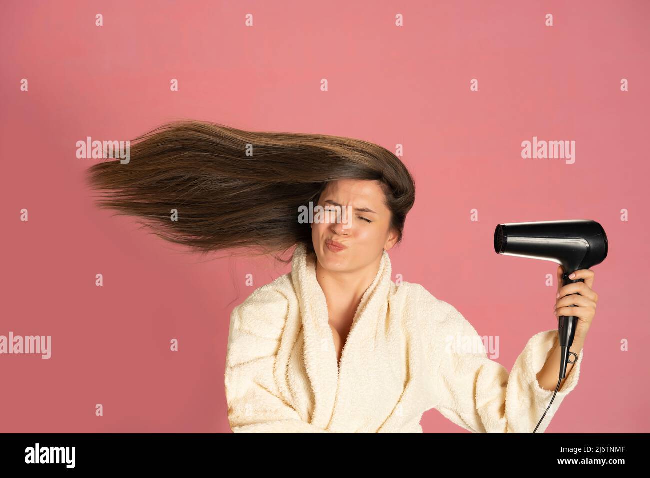 Funny woman drying her long hair with electric fan on a pink background ...