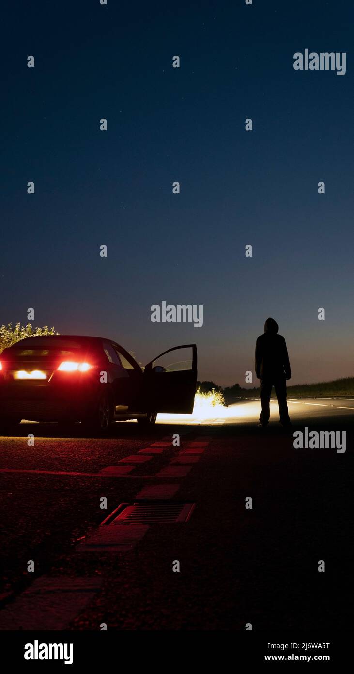 A mysterious hooded figure standing next to a parked car at night. On a ...