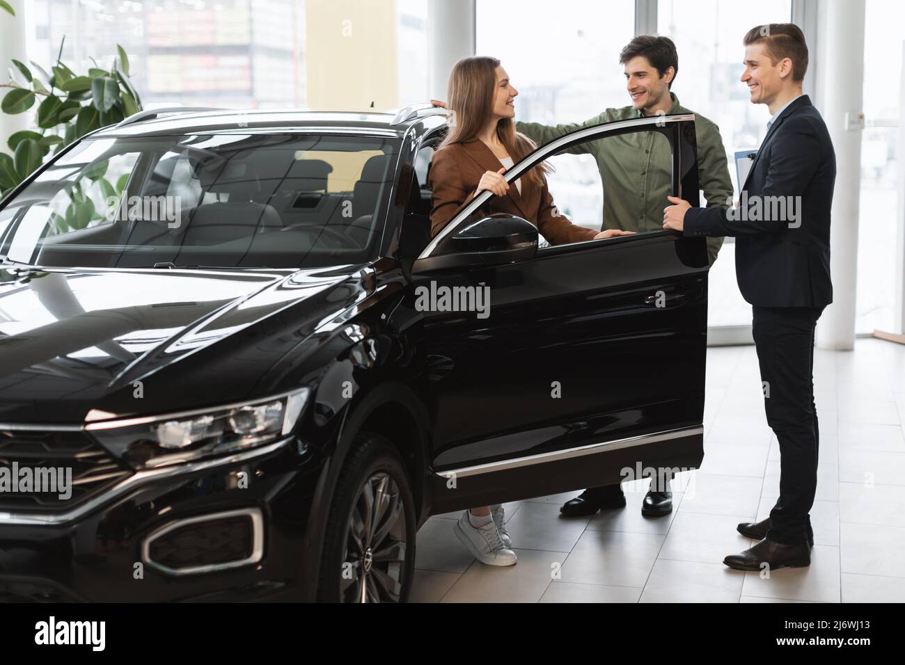 Cheery young couple speaking about buying new car with auto salesman at ...