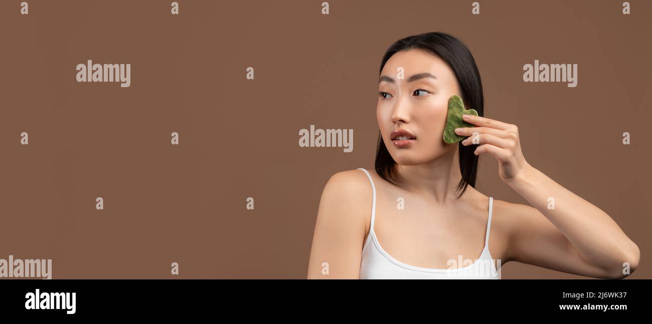 Sensual korean woman doing skin care routine, scrapping, massage facial skin with jade gua sha ...