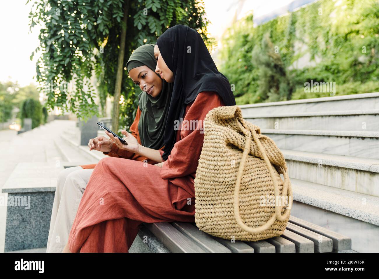 Multiracial muslim women using mobile phone while sitting on bench ...