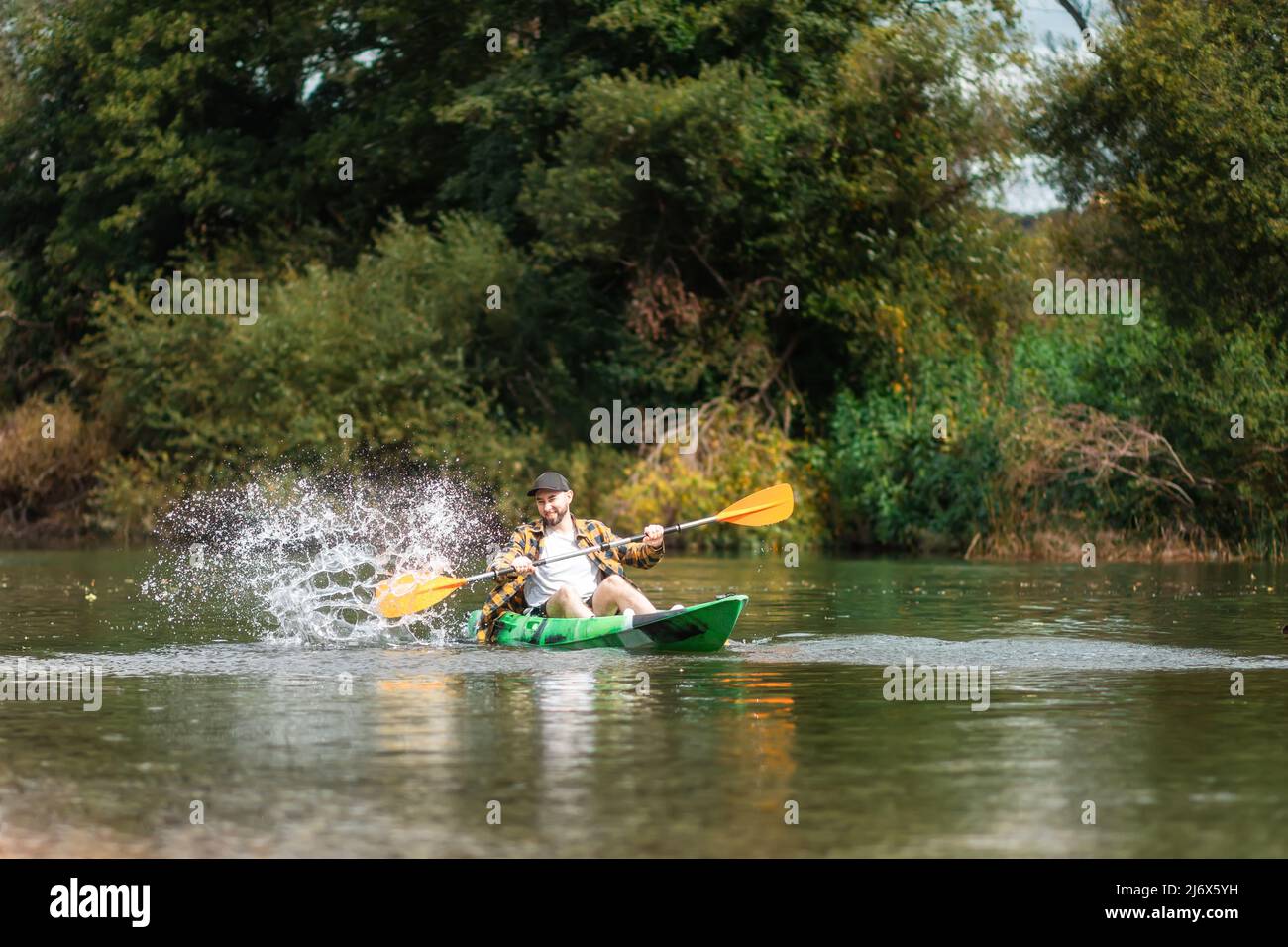 Happy bearded man in shirt and cap kayaking at the river with splashes ...