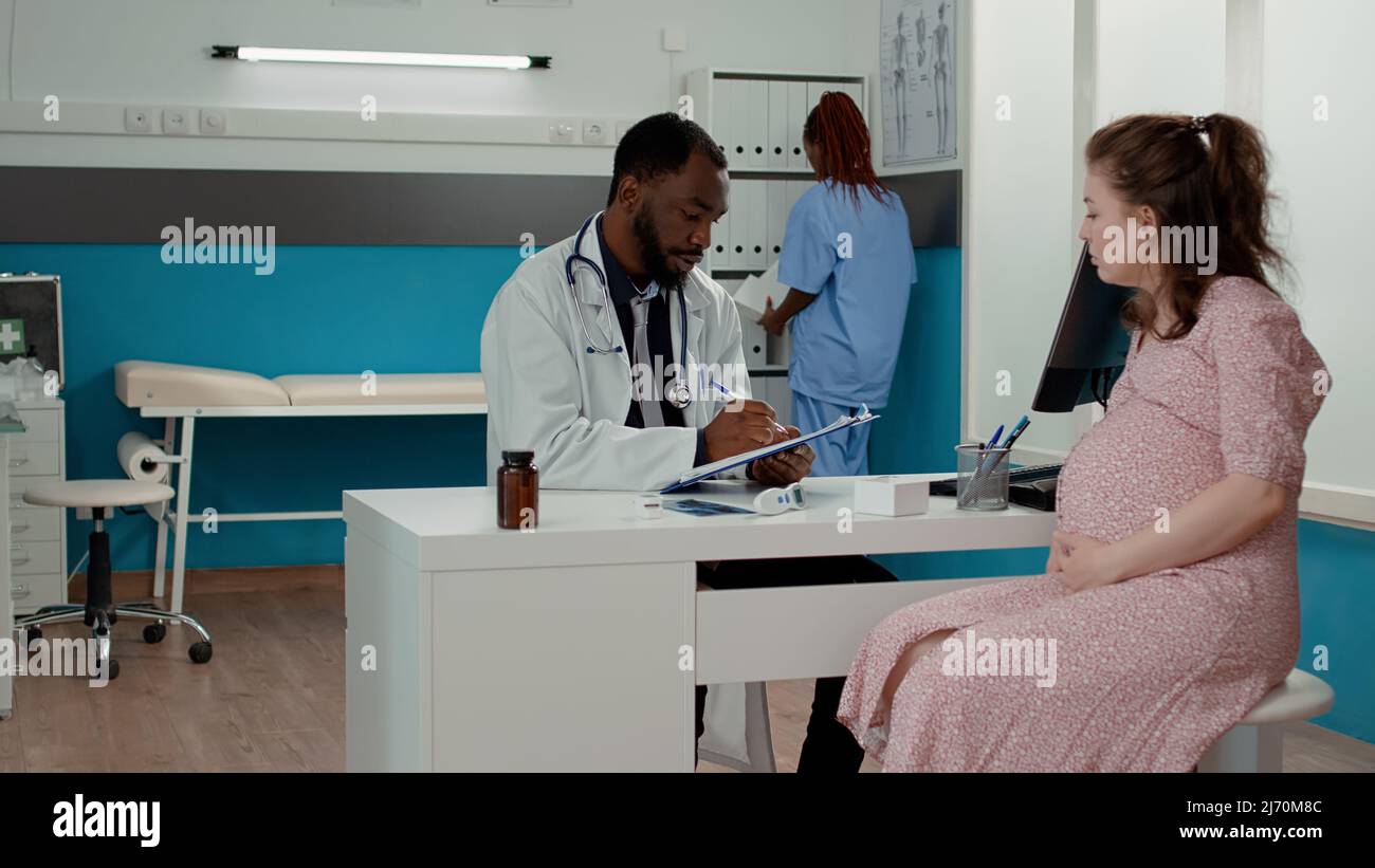 Physician consulting pregnant person and taking notes on checkup ...