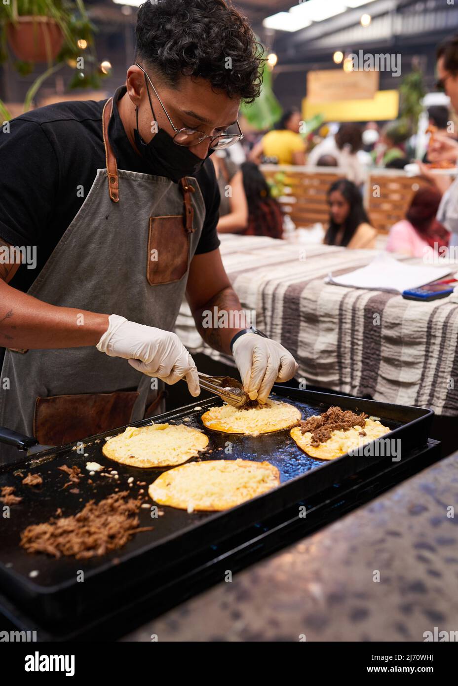 A chef spoons pulled beef onto tacos on the grill at a busy food market ...