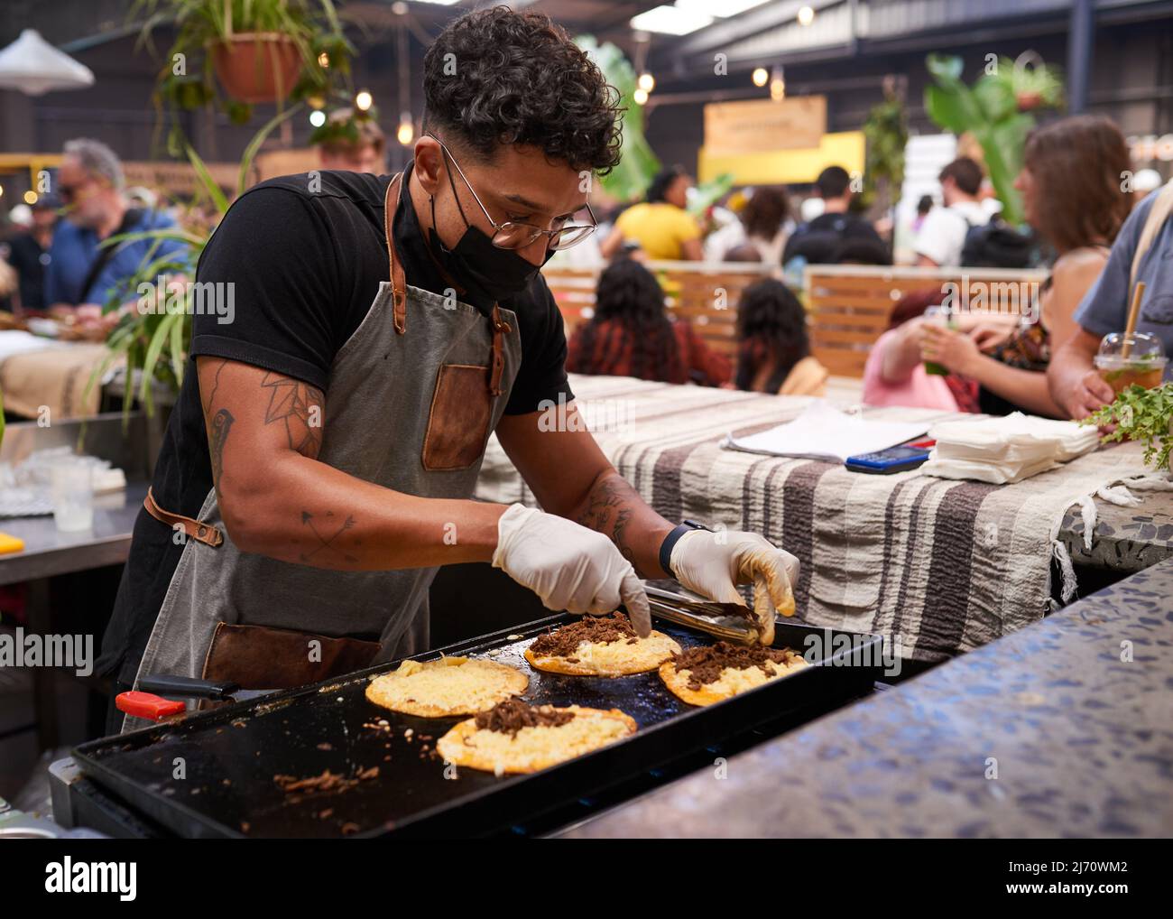 A multi-ethnic chef spoons meet onto tacos at a busy indoor food market ...