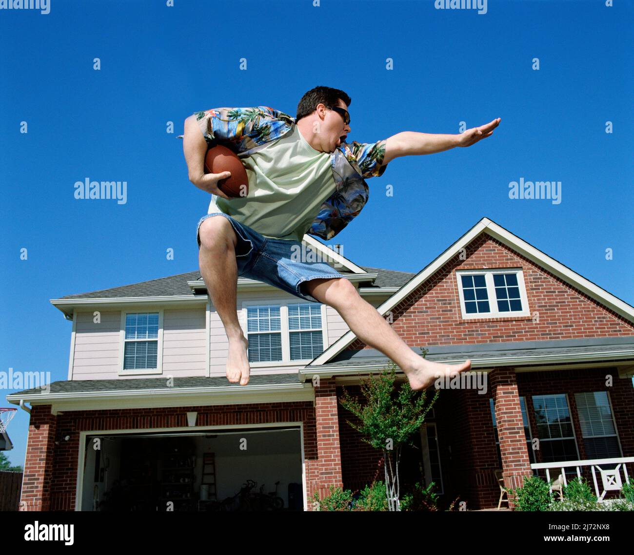 Middle aged man holding a football jumping in front of home, striking a ...
