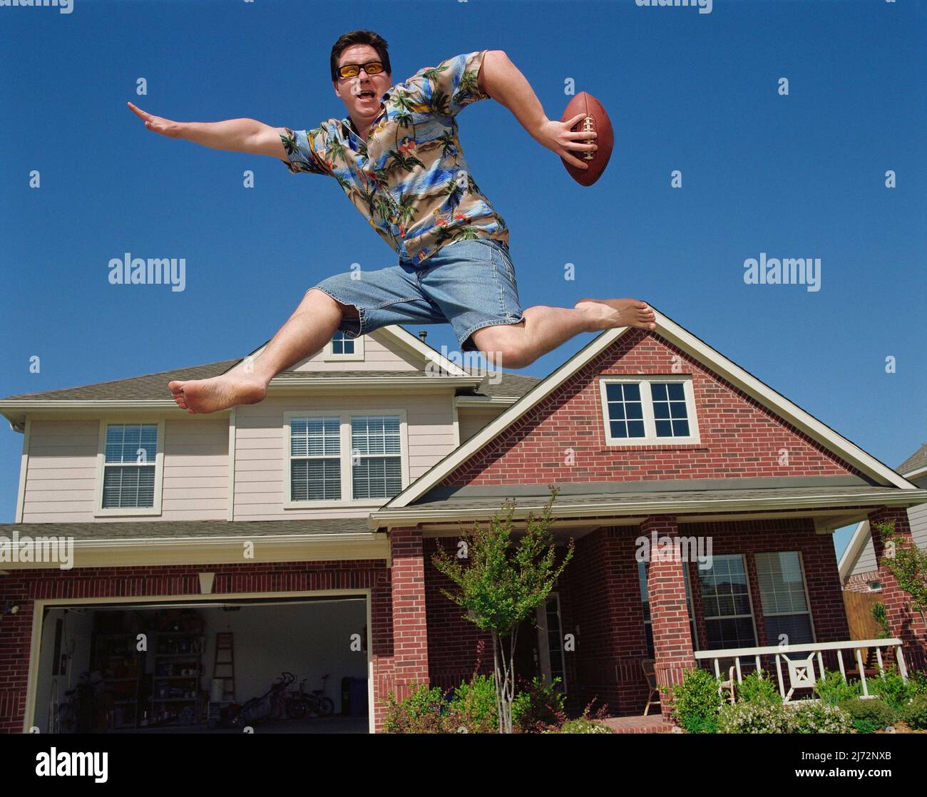 Middle aged man holding a football jumping in front of home, striking a ...