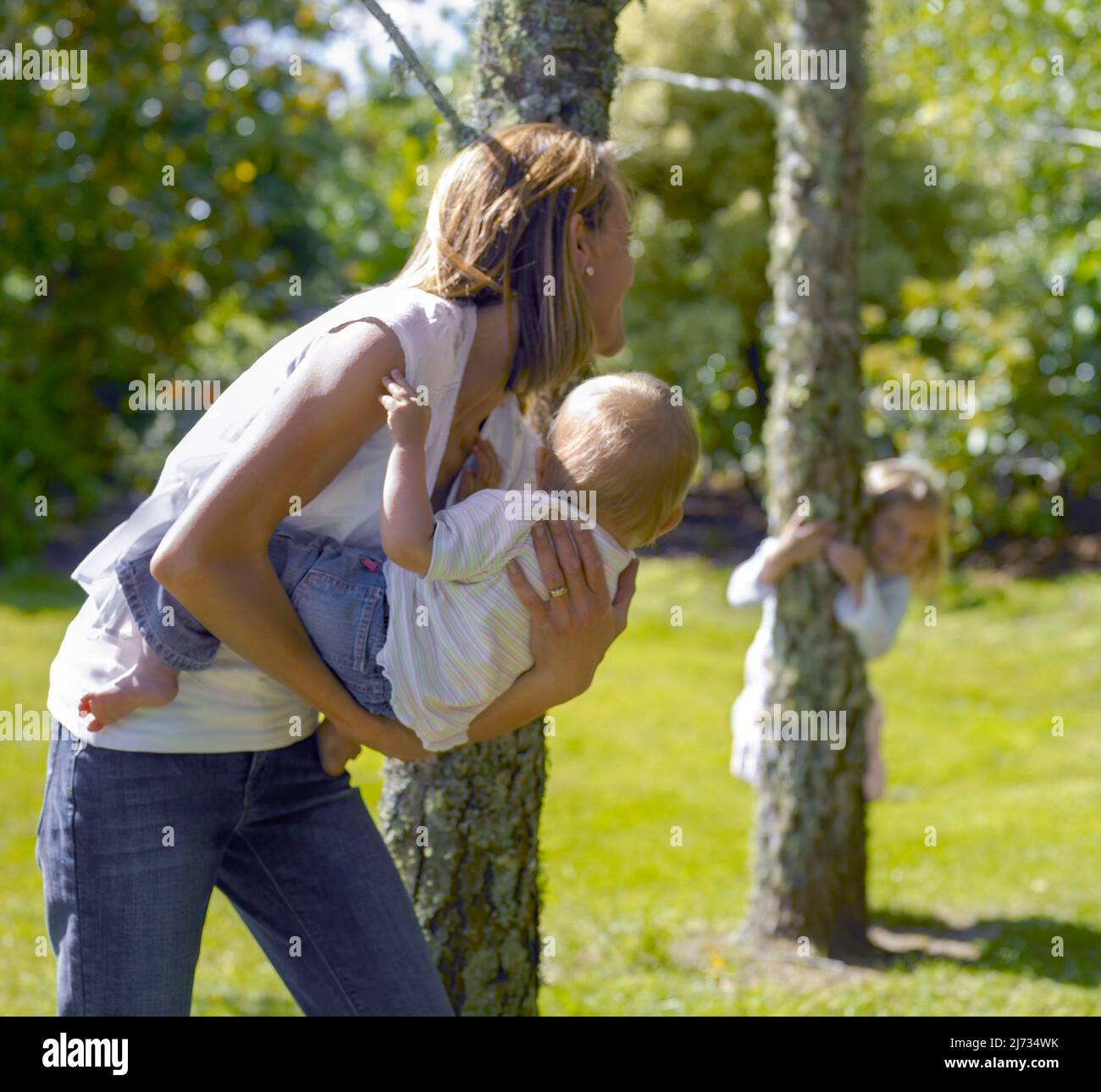 Mother holding baby and hiding behind tree from little girl paying hide ...