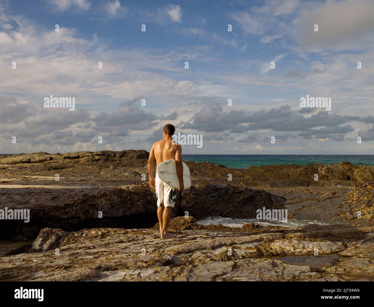 Back view of surfer carrying surfboard walking across rocky surface ...