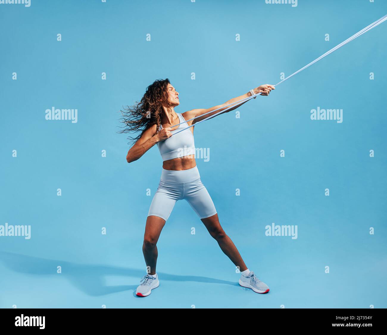 Muscular sportswoman exercising with resistance band on blue background ...