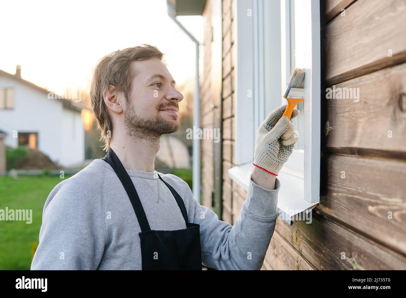 Young smiling man in apron painting window of his house with paint ...