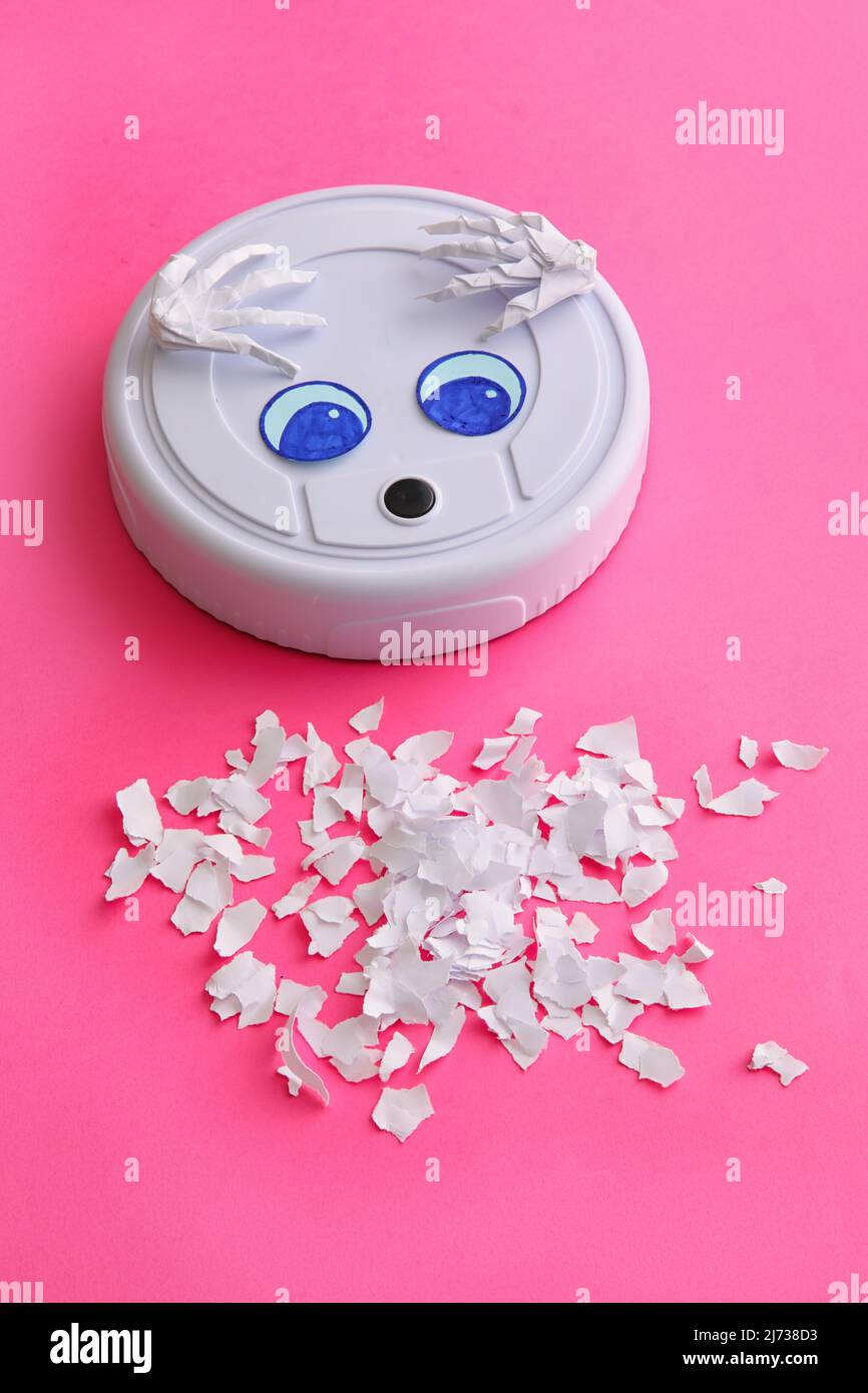 Modern robot vacuum cleaner with drawn eyes and paper pieces on pink ...