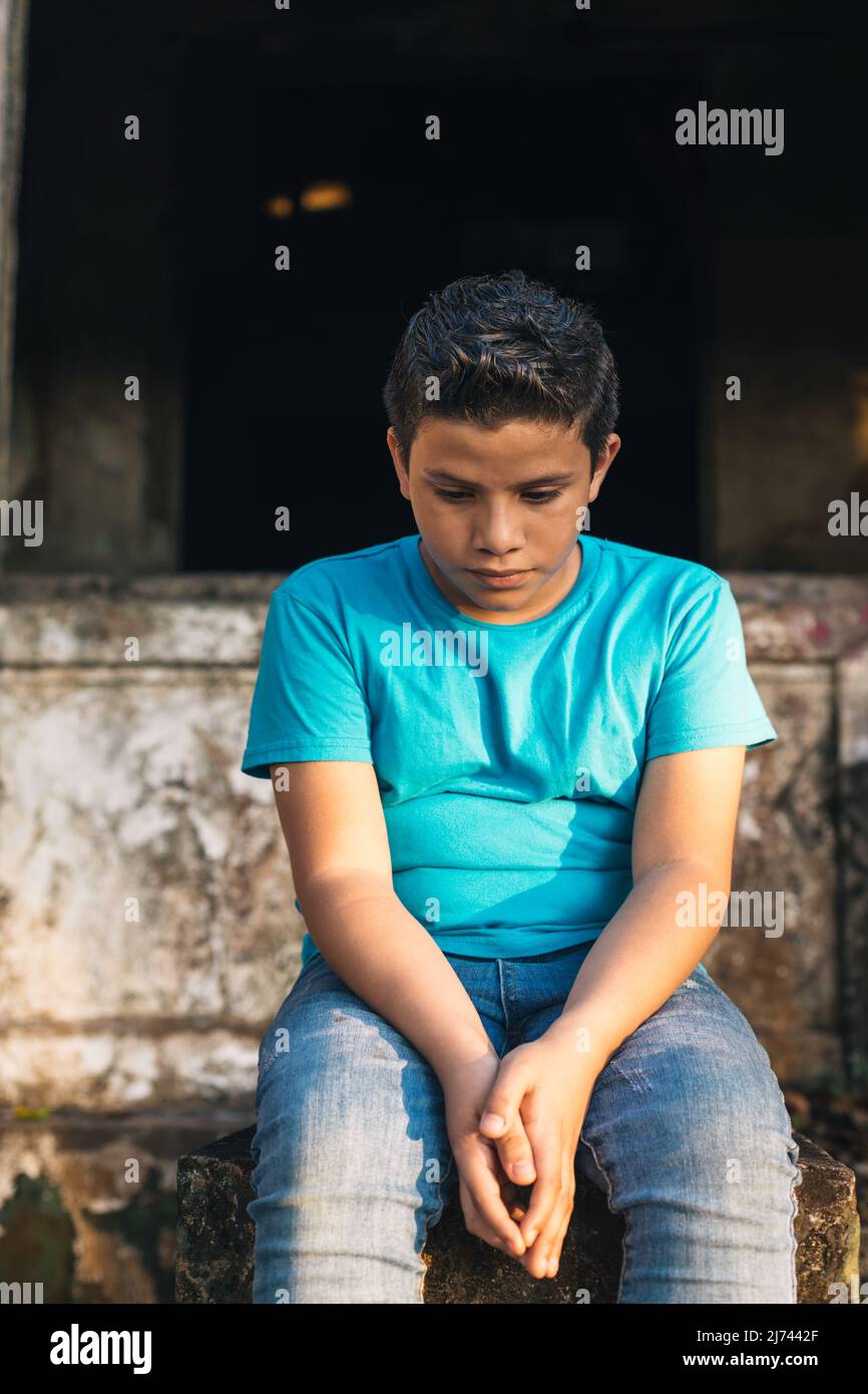 Hispanic boy sitting on some ruins looking at the ground, with a sad ...