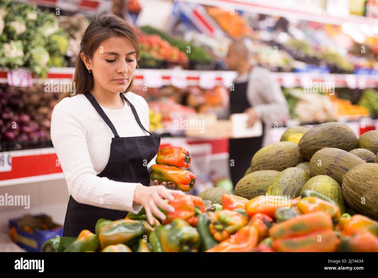 Female supermarket employee in apron lays fresh bell peppers on counter ...