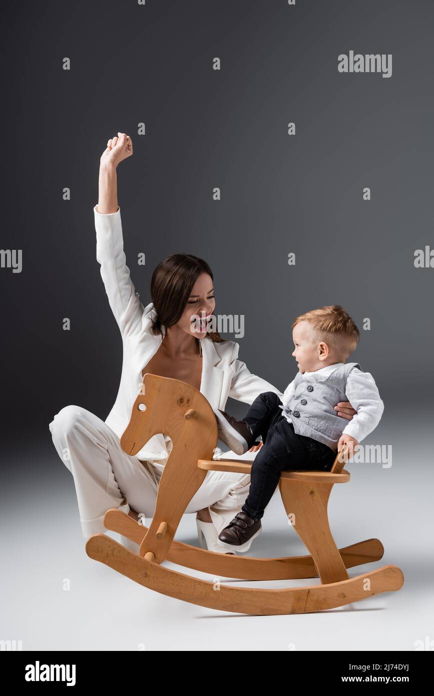 excited mother showing triumph gesture near little son on rocking horse ...