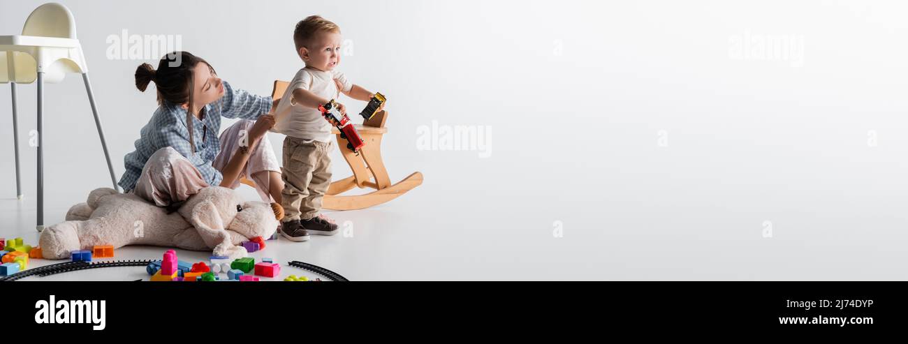 toddler boy riding rocking horse near young mother and toys on grey ...