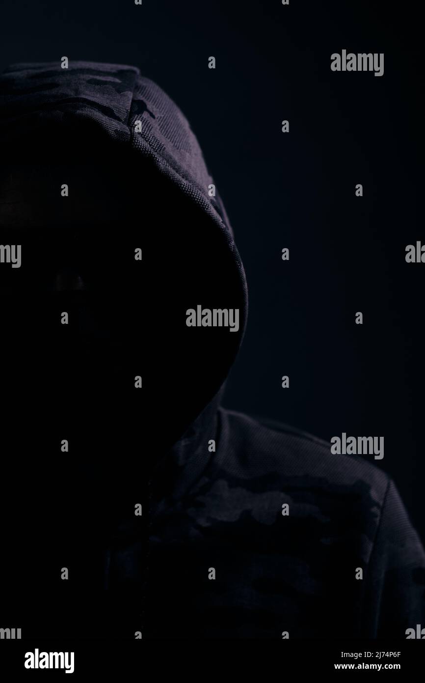 Man in dark camouflage hoodie with covered face dark background, hacker ...