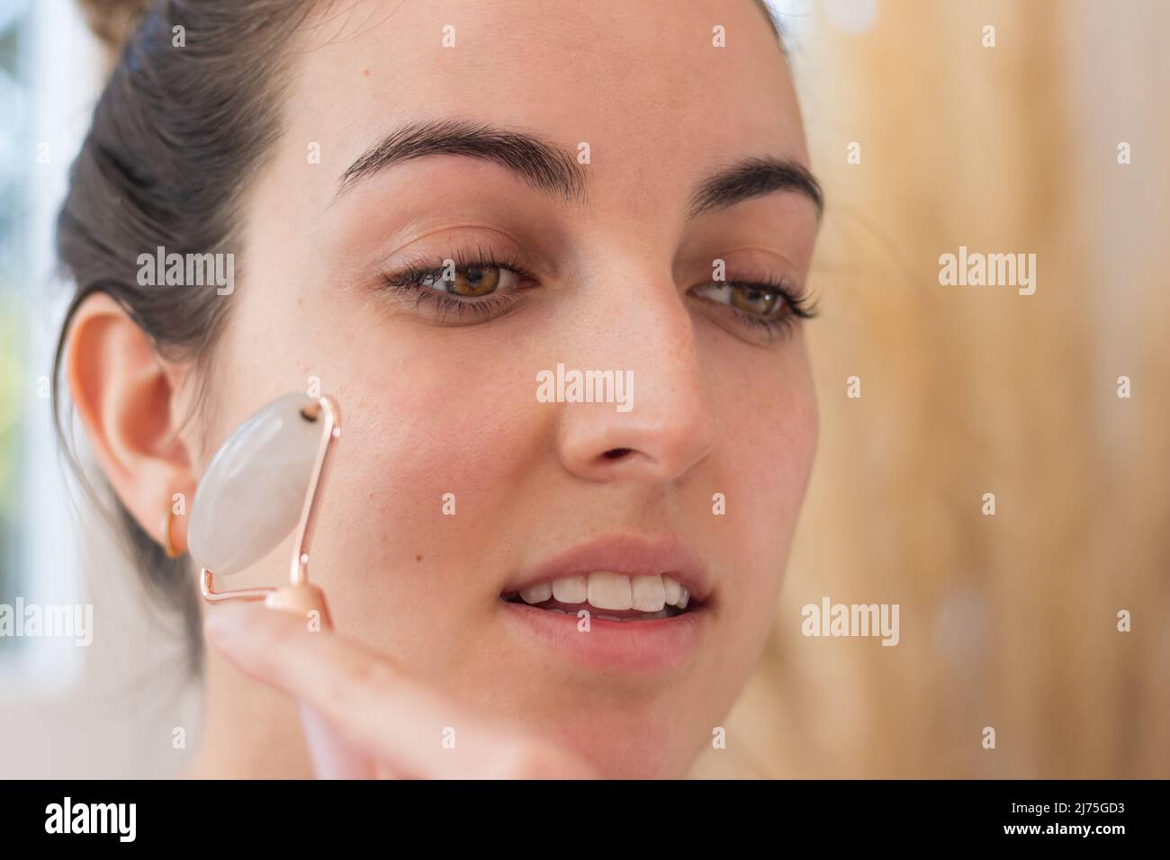 Smiling and happy woman using face roller on her cheek while doing her ...