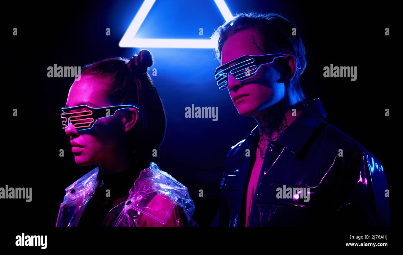 Young cyber couple in vinyl jackets and LED goggles standing in dark ...