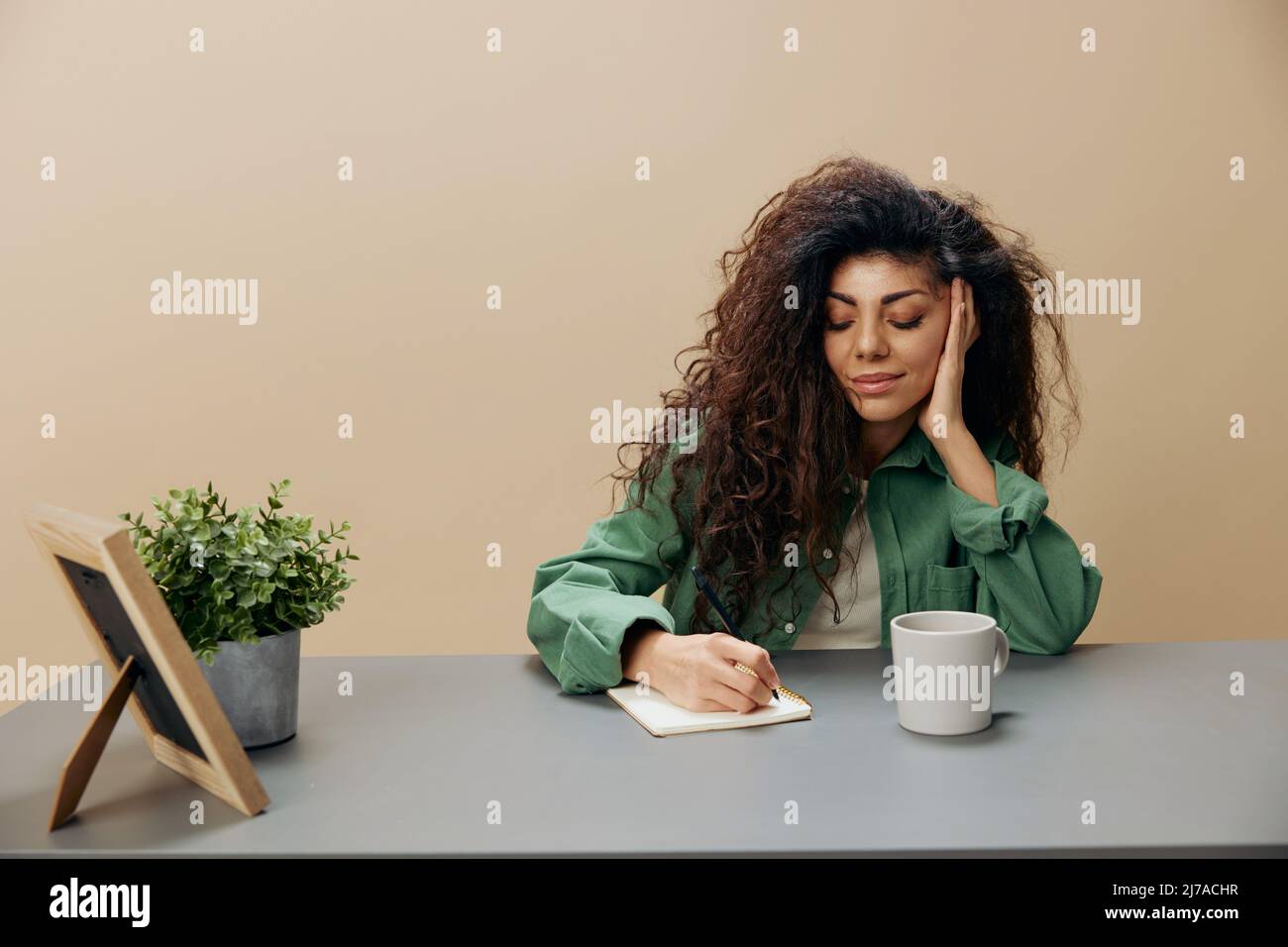 Enjoyed smiling tanned curly Latin lady write letter sit at the table ...