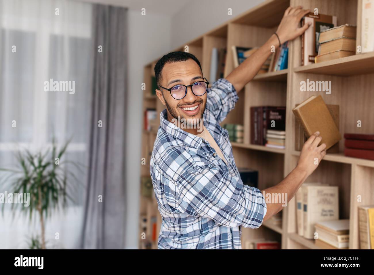 Home library. Happy arab man taking book from bookshelf, searching and ...