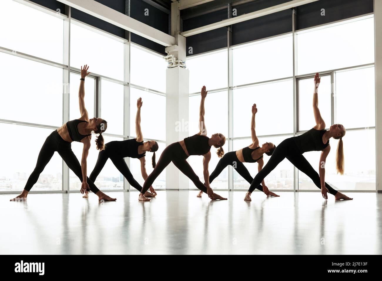Group of young women doing triangle pose against panoramic window while ...