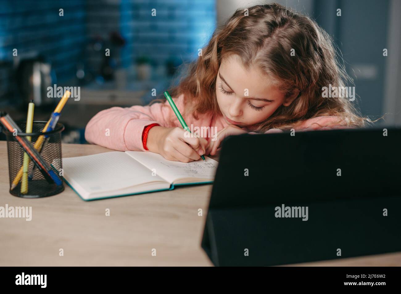 Schoolgirl writing note by pencil sitting in front of computer tablet ...