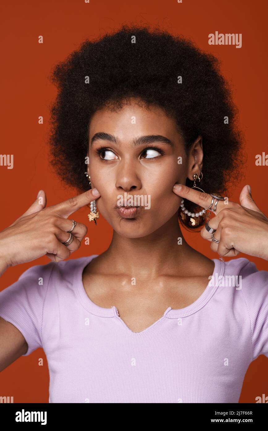 Black young woman wearing rings pointing fingers at her cheeks isolated ...