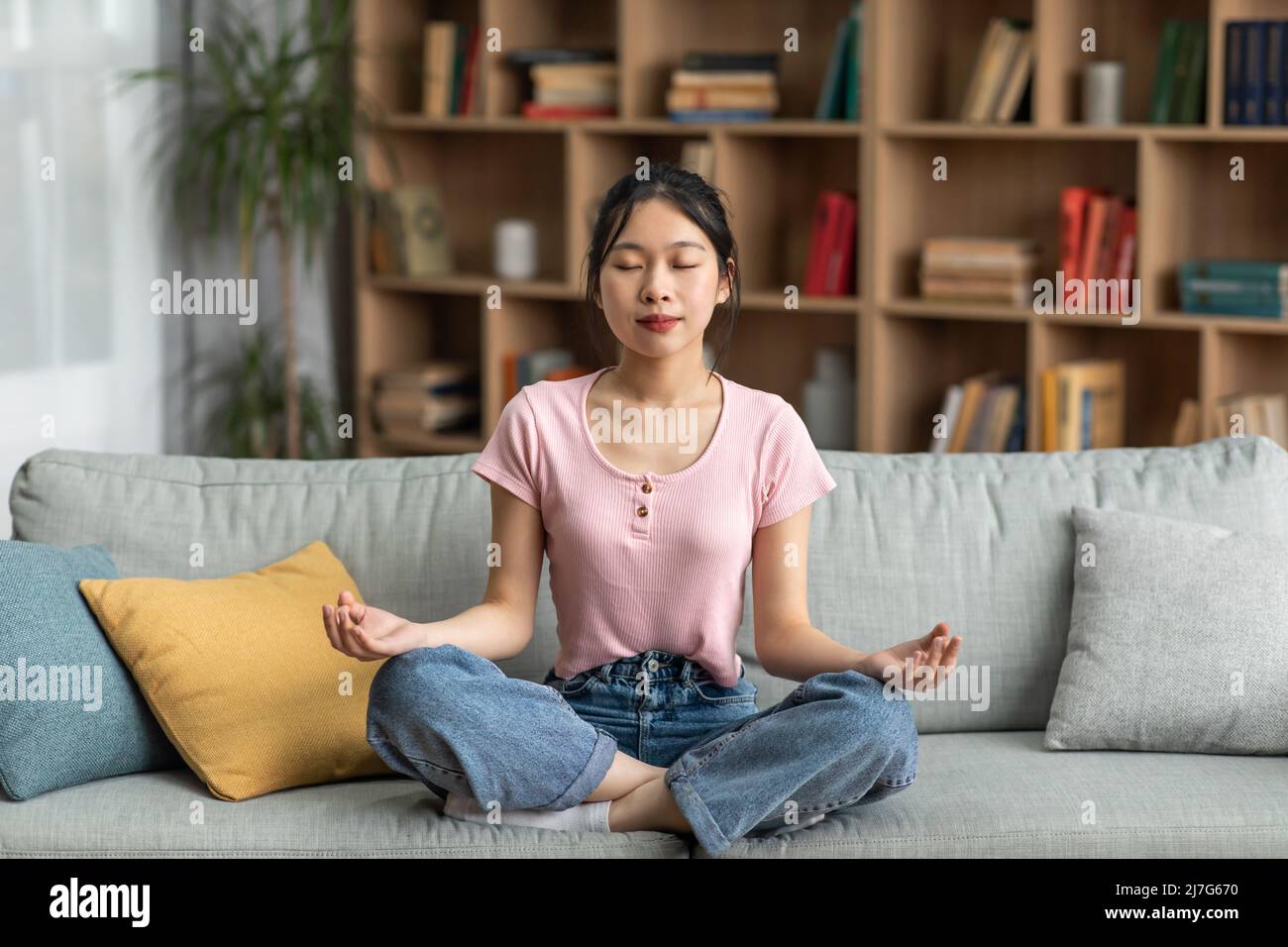 Calm asian lady sitting on comfy sofa and meditating with closed eyes ...