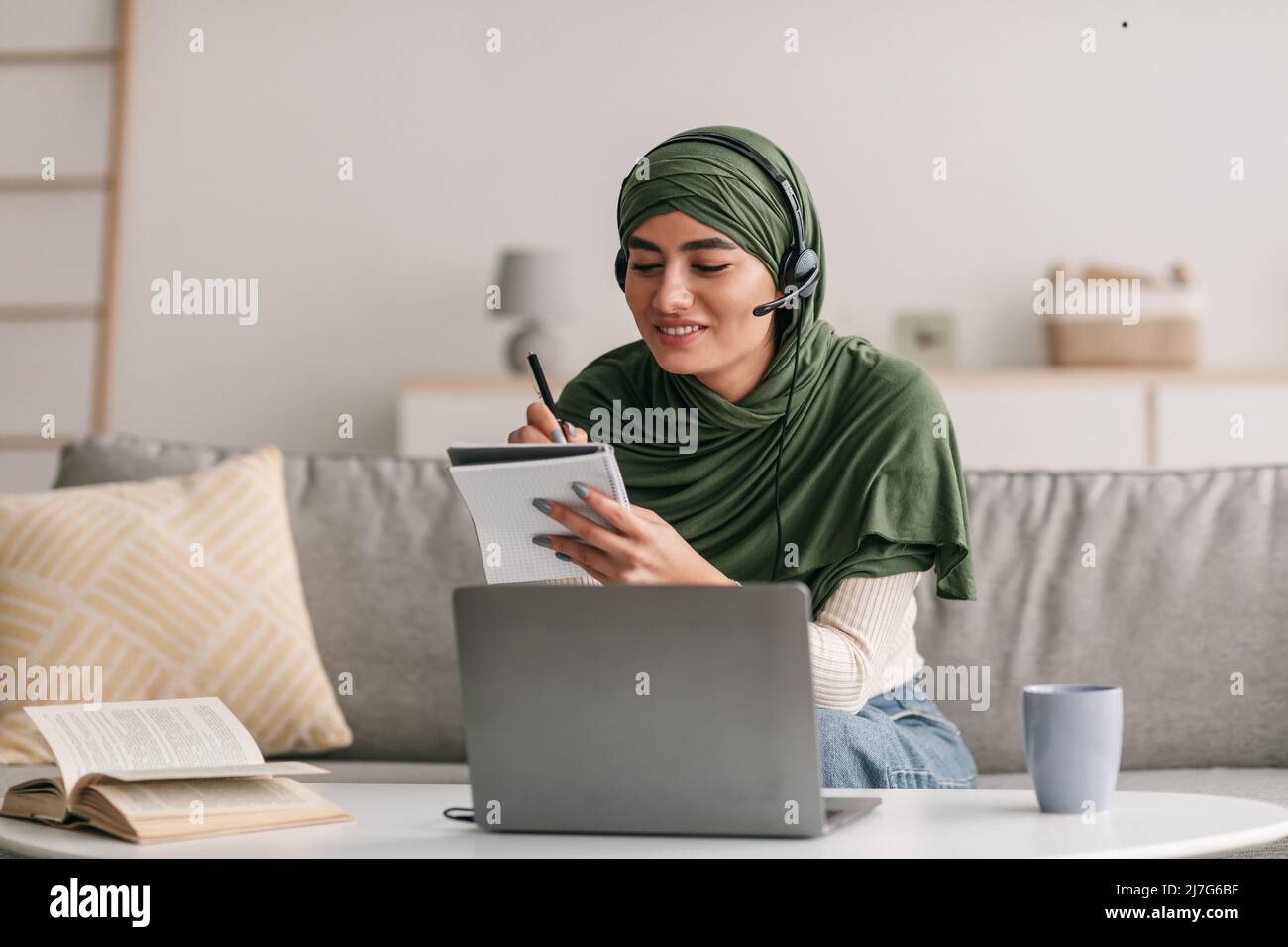 Cheerful Arab woman in hijab using laptop, wearing headphones, writing ...