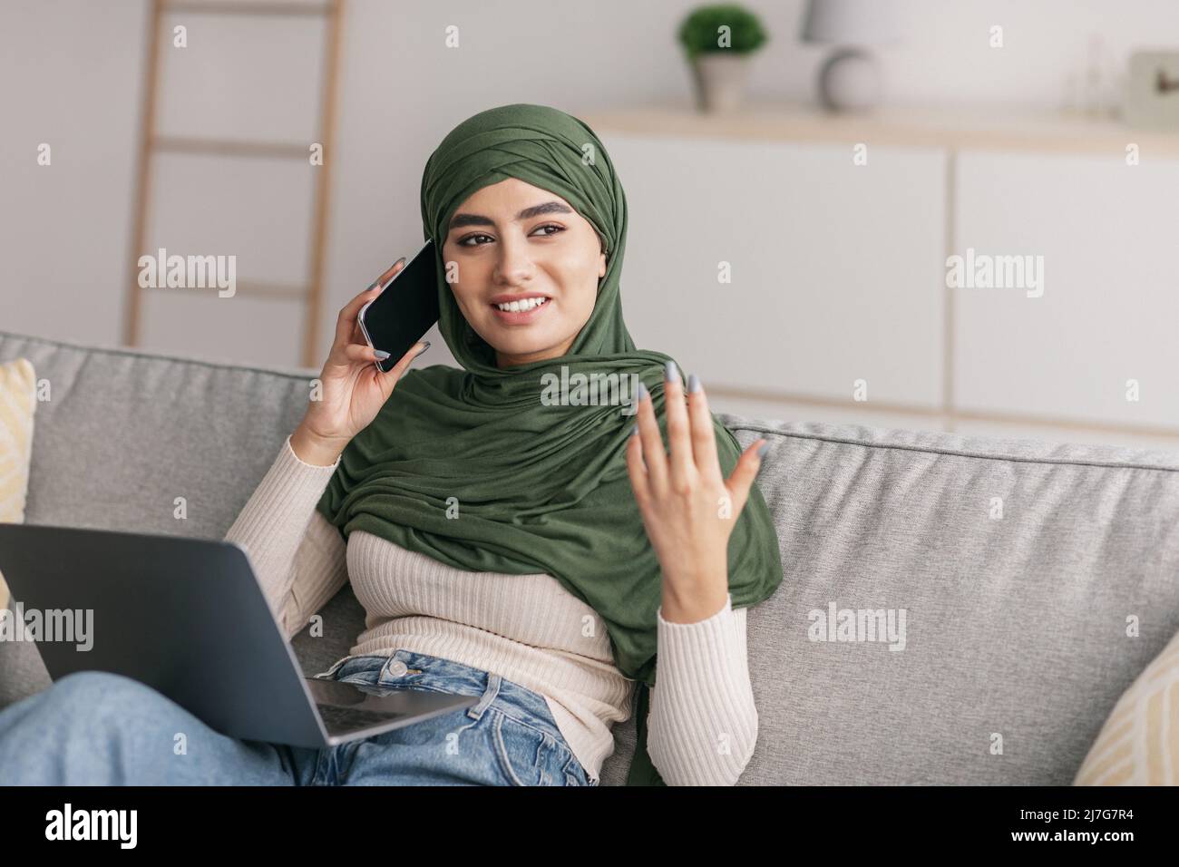 Positive young Arab woman in hijab using laptop and speaking on ...