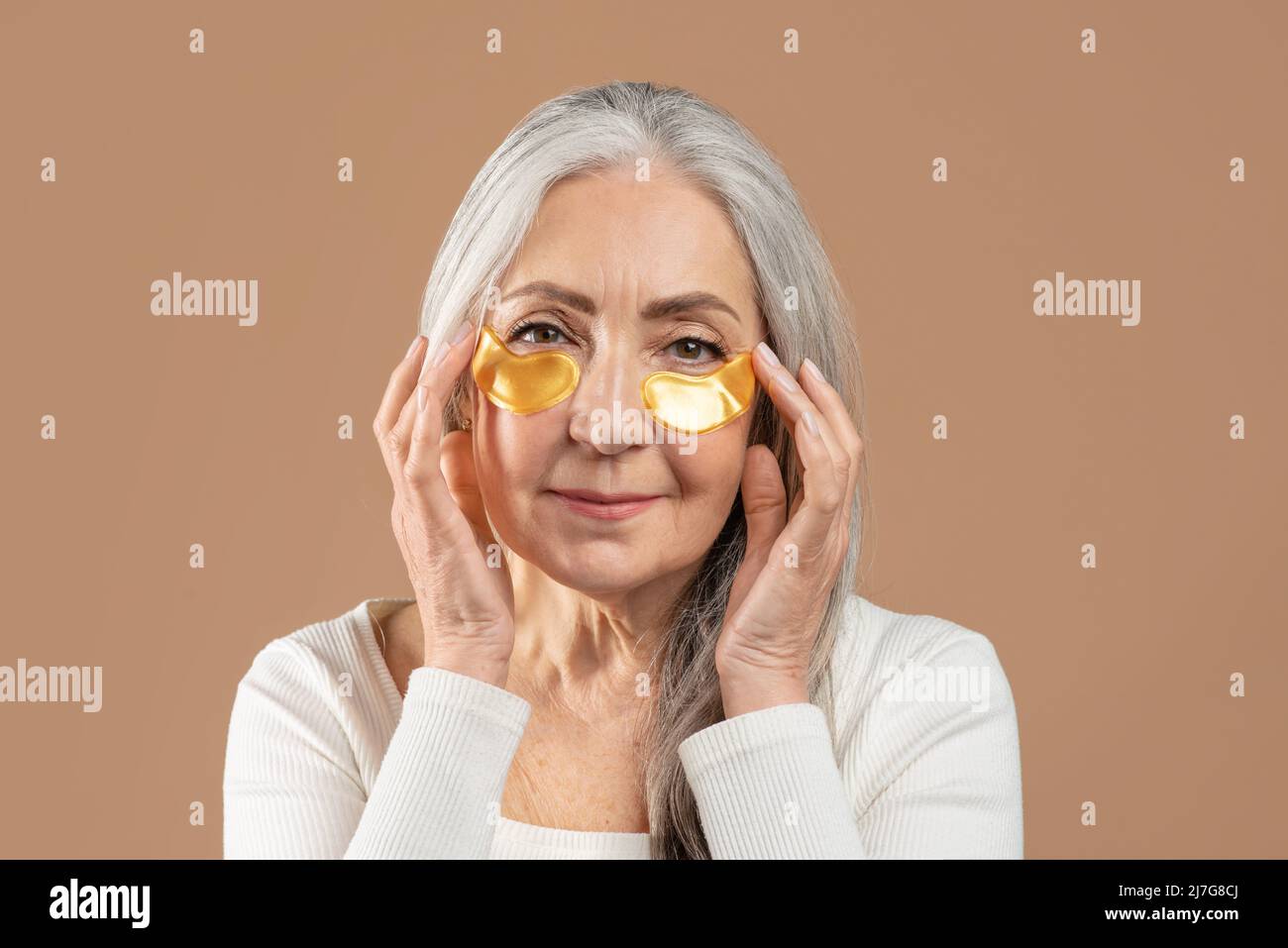 Feminine senior lady wearing cosmetic eye patches on brown studio ...