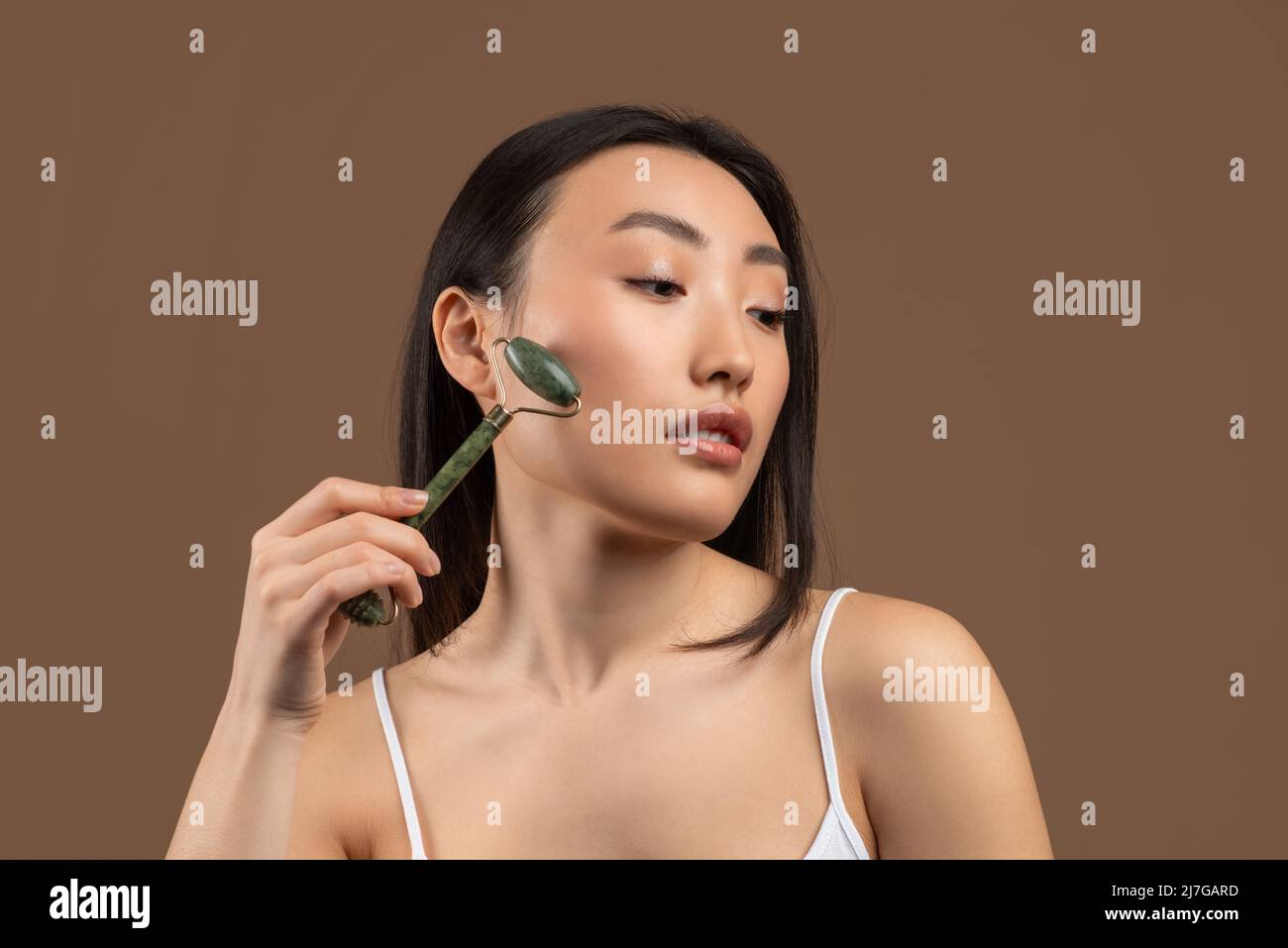 Young asian lady using greenstone jade facial roller, looking aside ...