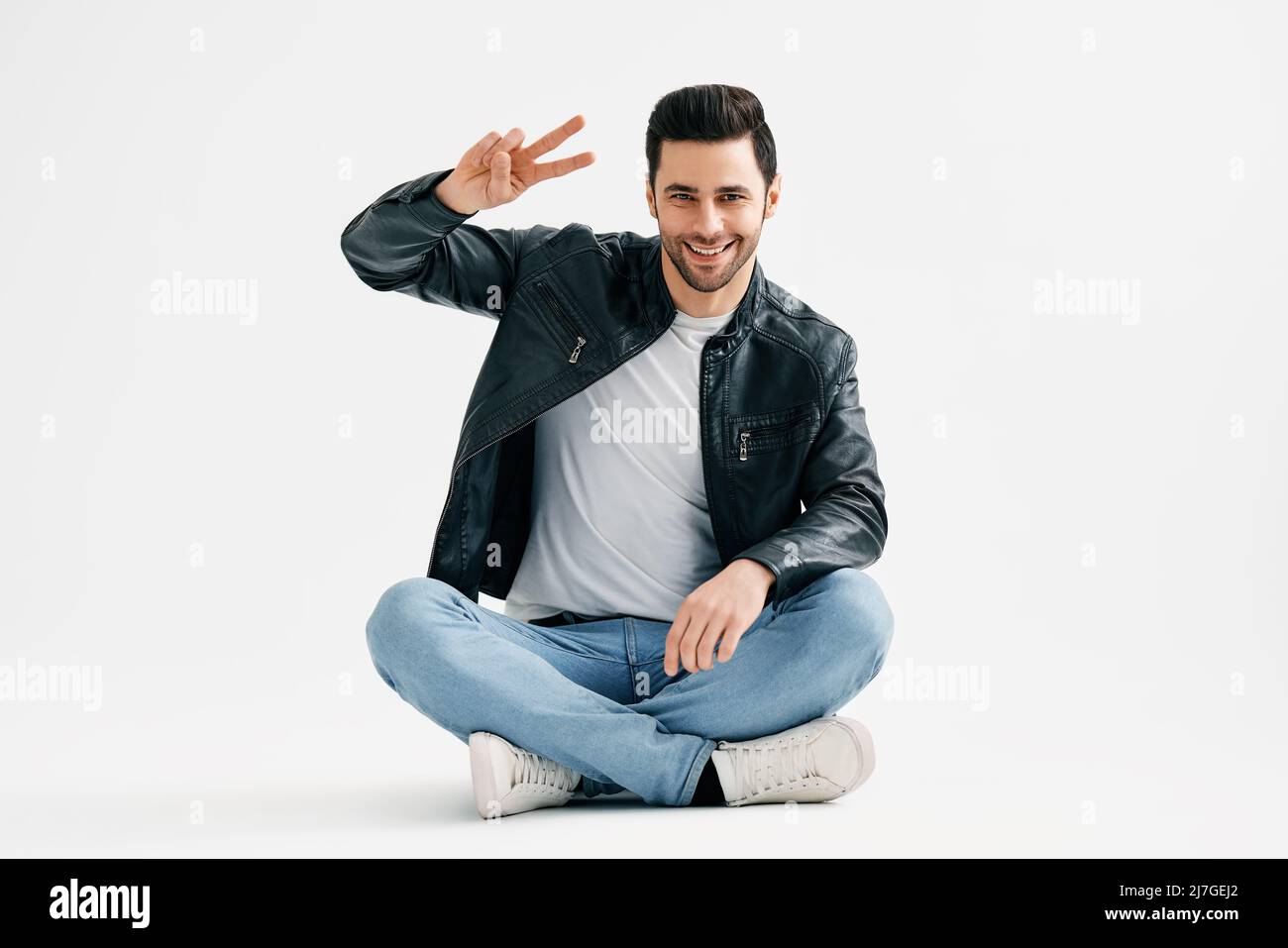 Positive cheerful young man doing victory sign sitting with legs ...