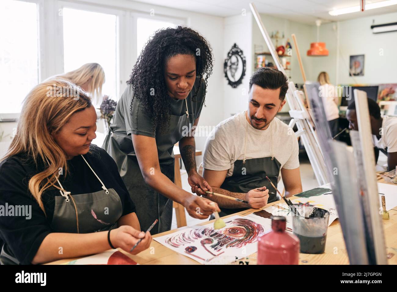 Mature female instructor teaching painting to young students at desk in ...